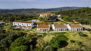 Um conjunto de edifícios brancos com telhados marrons, cercado por vegetação e colinas ao fundo.