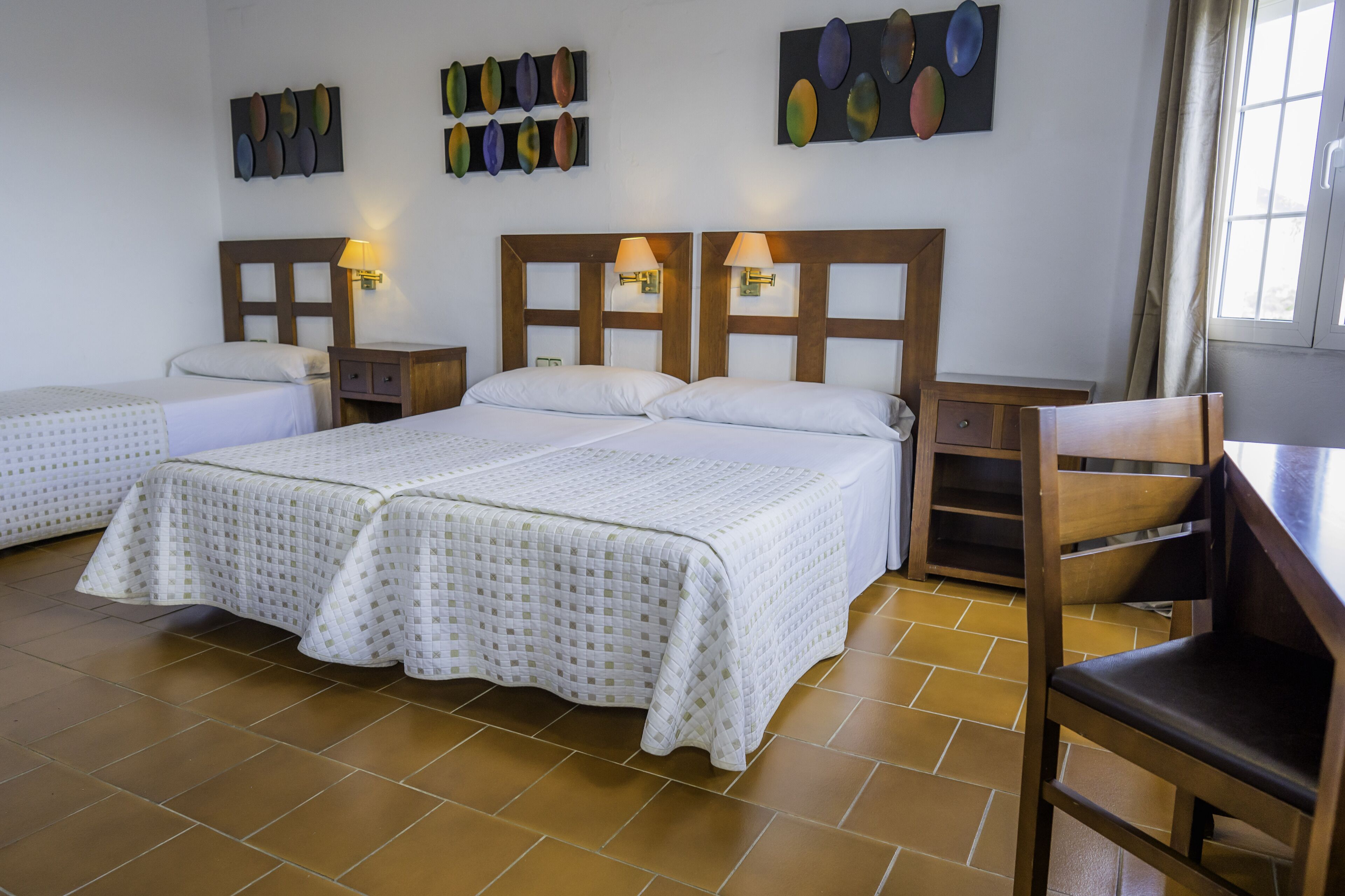 Quarto de hotel com duas camas, cabeceira de madeira, mesa de cabeceira, uma cadeira e uma janela com cortinas.
