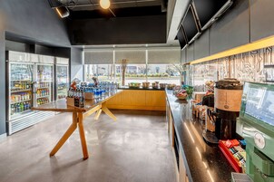 Interior moderno de café com uma mesa central de madeira, balcão refrigerado para bebidas e uma máquina de café.