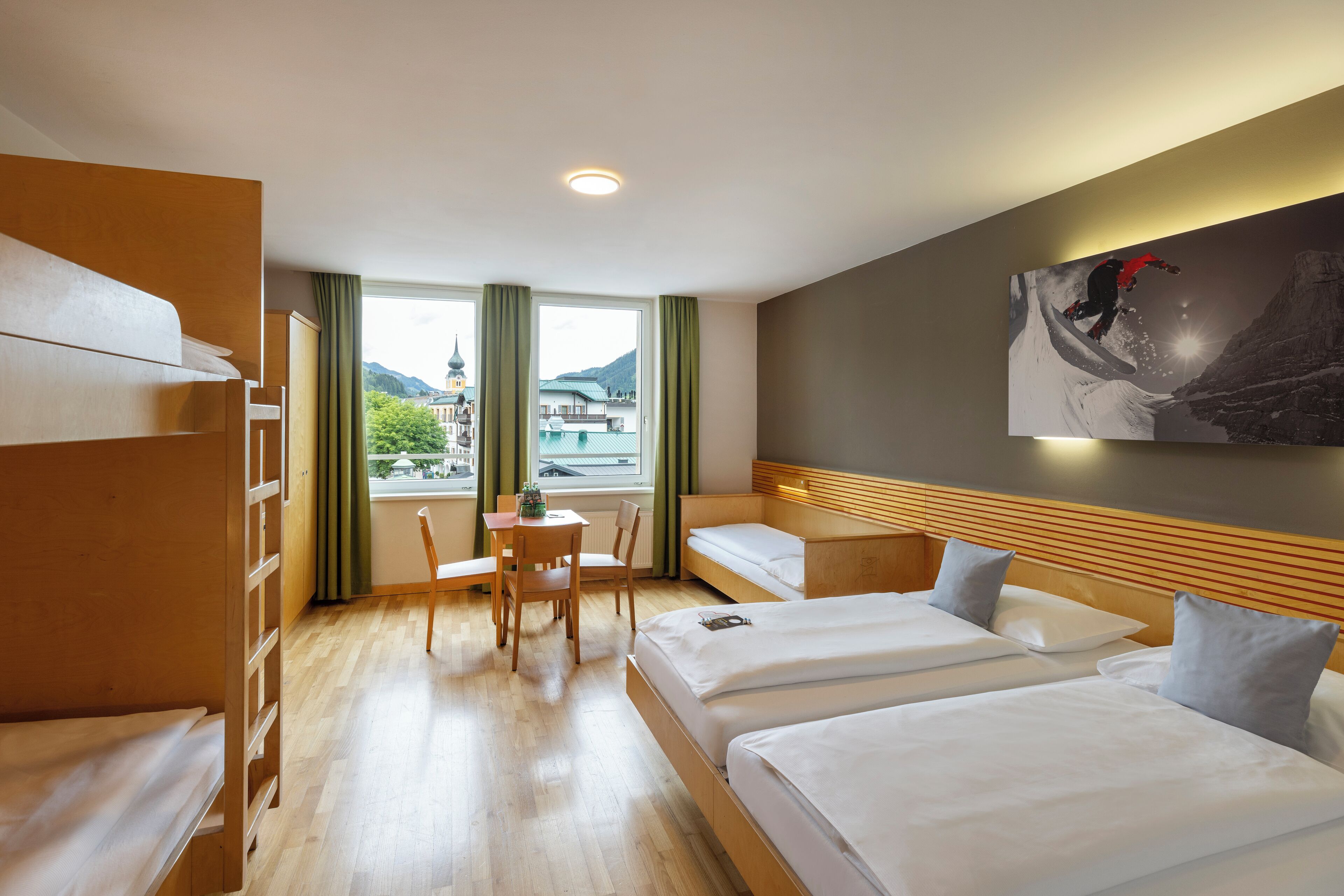 Quarto de hotel com duas camas, uma escrivaninha, uma cadeira e uma janela grande com vista para edifícios e montanhas.