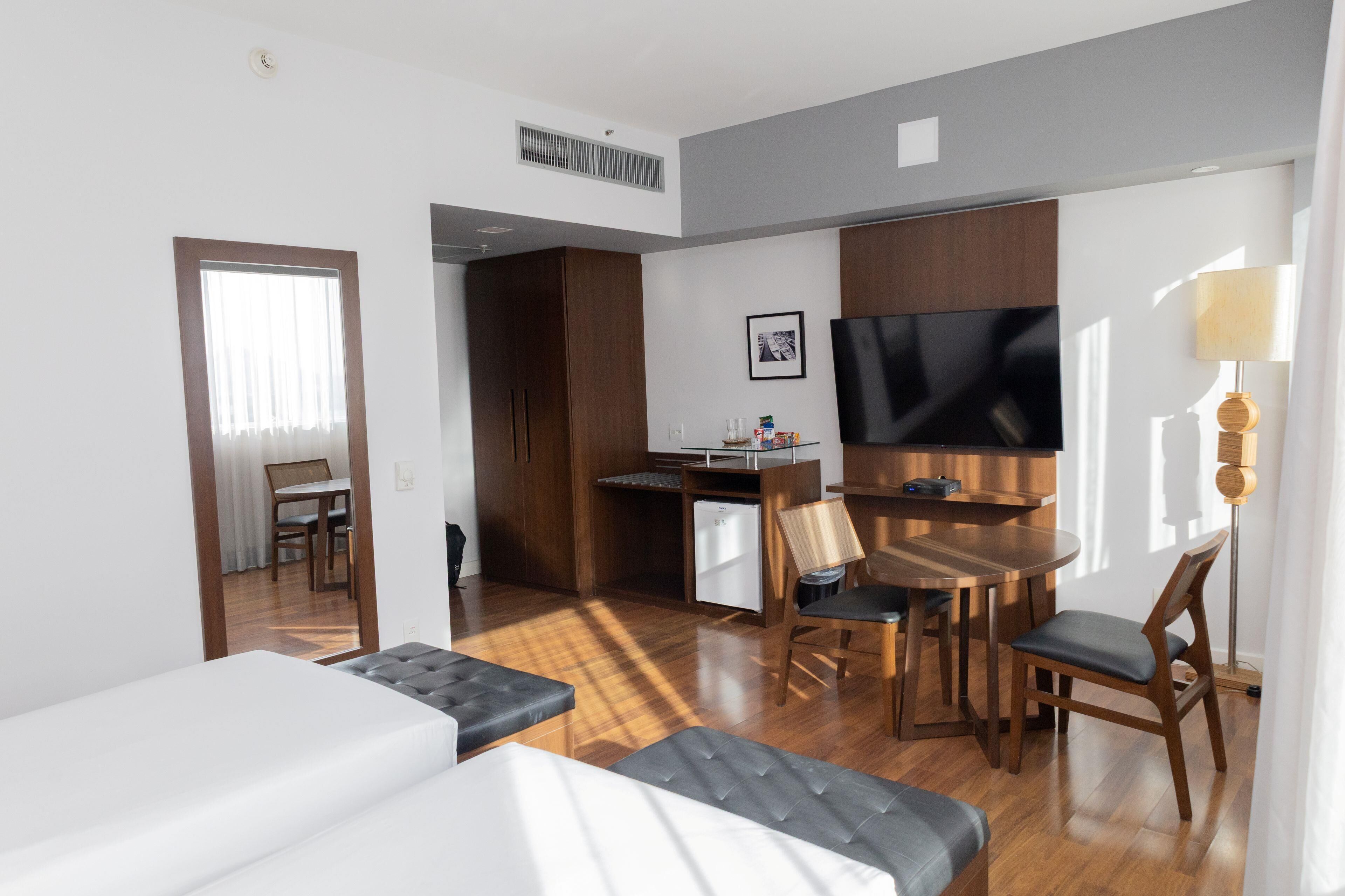 Quarto de hotel com cama, escrivaninha, cadeira, televisão e luminária.