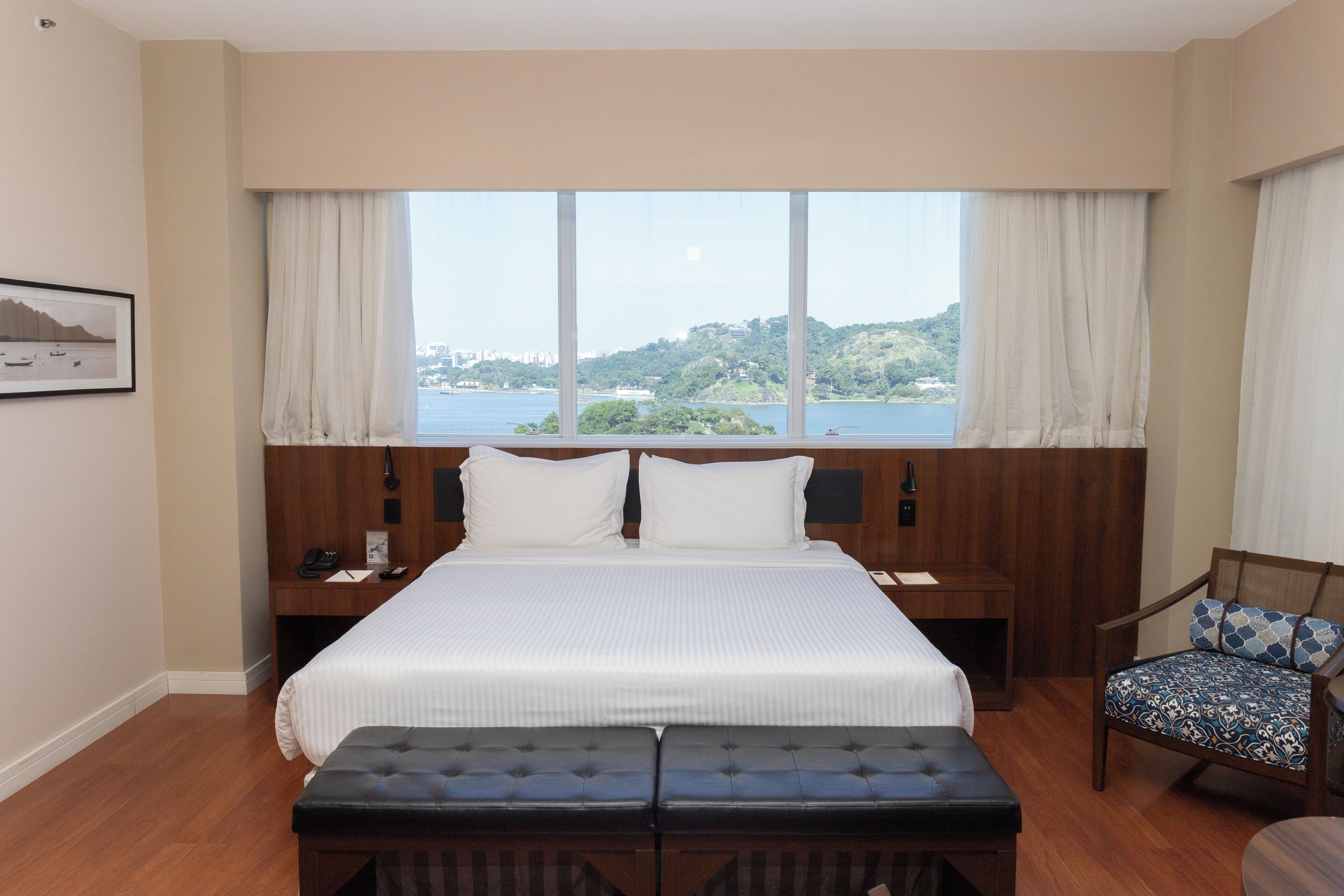 Quarto de hotel com uma cama grande, cabeceira de madeira, vista para um corpo de água, uma cadeira e um banco.