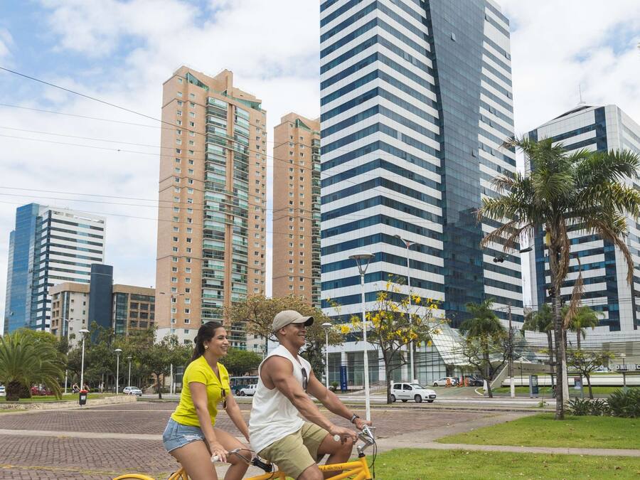 Um homem e uma mulher pedalando uma bicicleta amarela em um caminho pavimentado, com edifícios altos ao fundo.