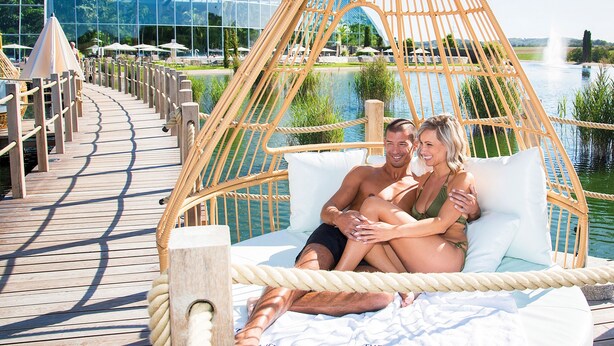 Um casal relaxando em uma cama de vime com vista para um edifício moderno e um corpo d'água.