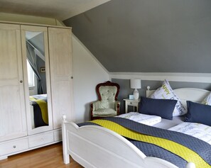 Um quarto com uma cama branca, um guarda-roupa, uma cadeira e uma mesa de cabeceira com um abajur e um relógio.