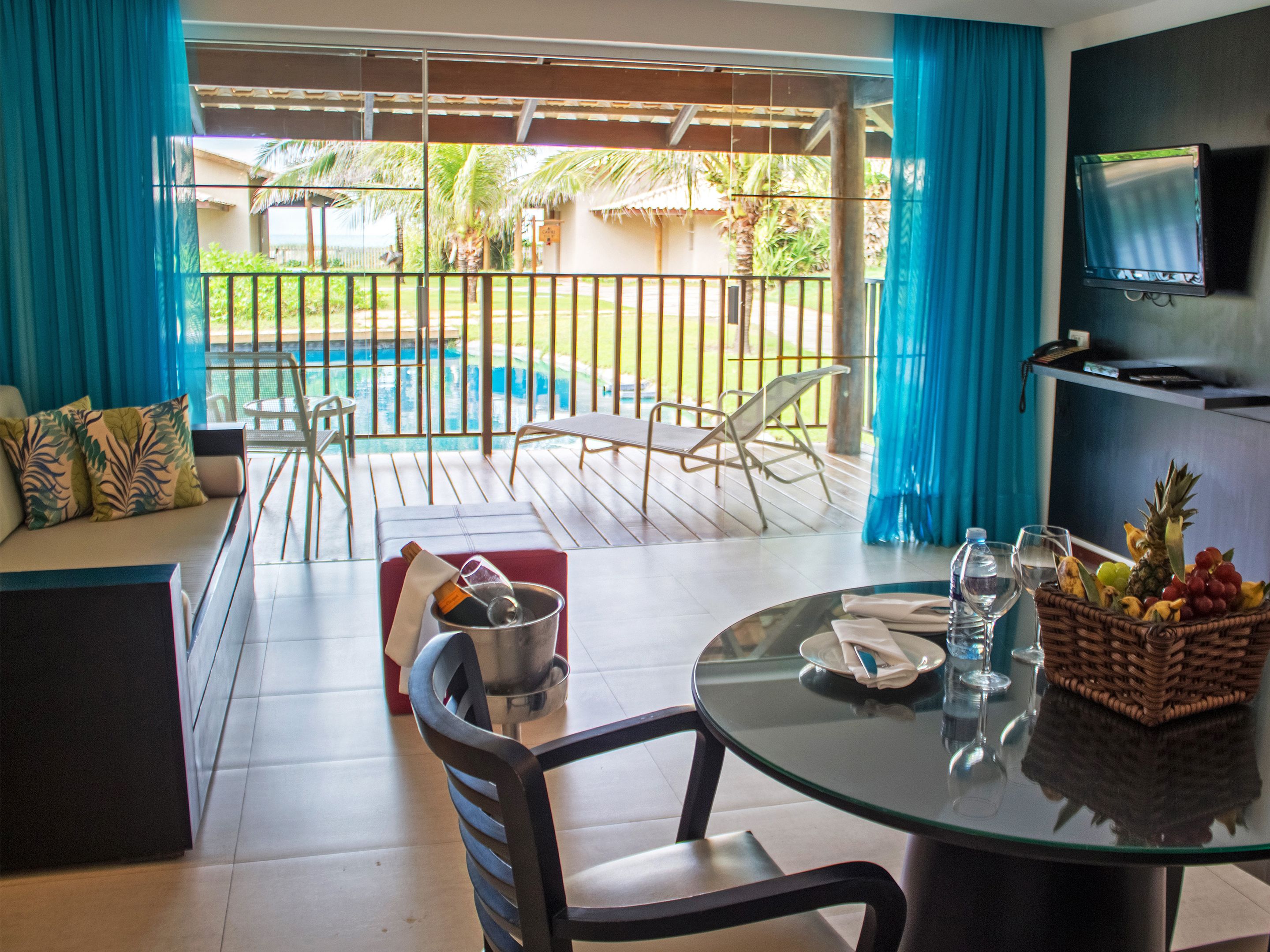 Quarto com mesa de vidro, cadeiras, televisão, vista para a piscina e cesta de frutas.