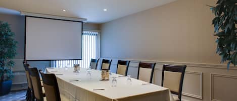 Uma sala de conferências com uma mesa comprida, cadeiras, uma tela de projetor e um vaso de plantas.