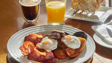 Prato de pequeno-almoço com ovos, bacon, salsichas e tomates. Há dois copos de bebidas e uma fatia de pão na mesa.