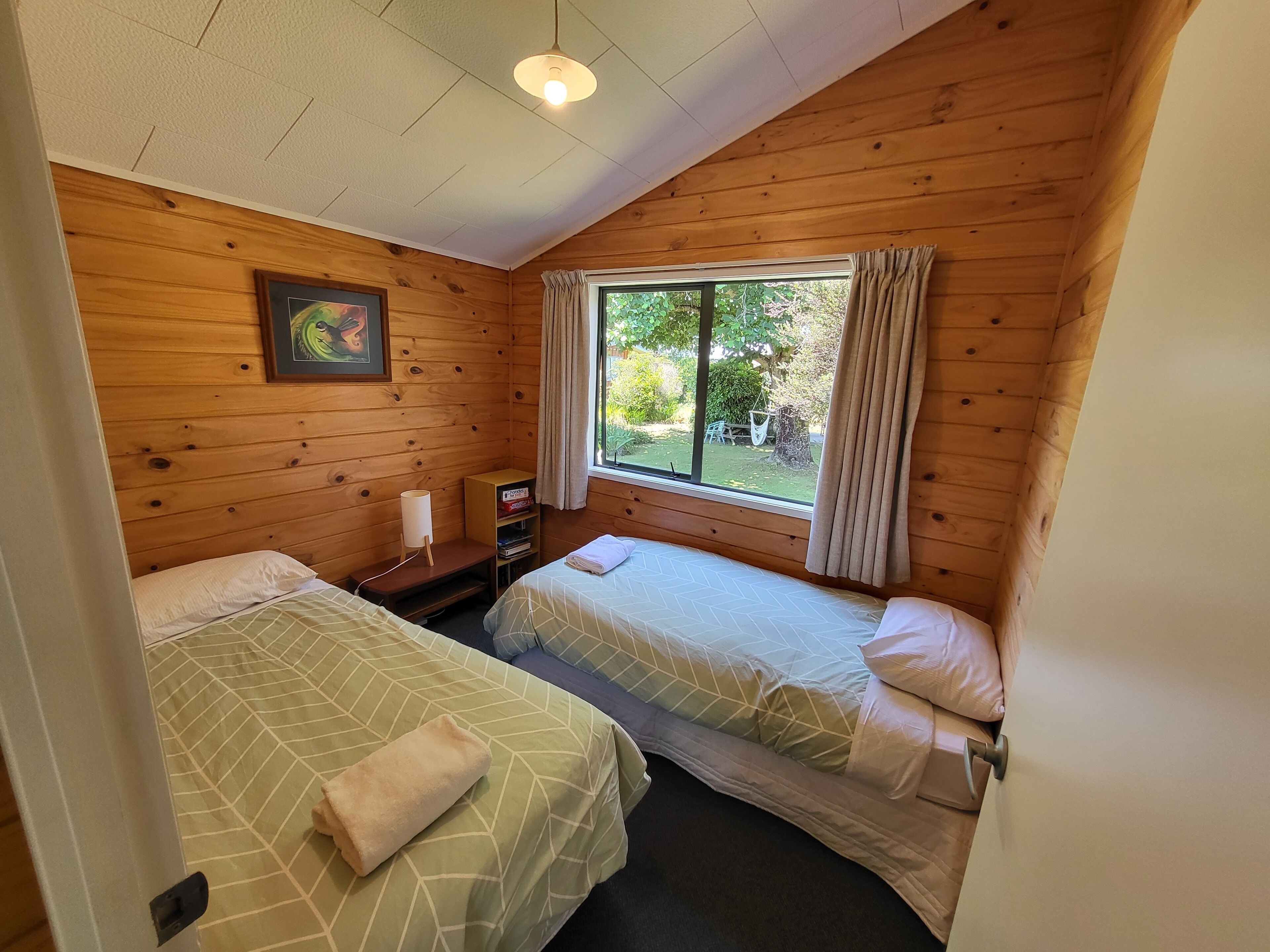 Quarto de cabana de madeira com duas camas, janela com cortinas, um quadro na parede e uma pequena mesa com abajur.
