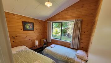 Quarto de cabana de madeira com duas camas, janela com cortinas, um quadro na parede e uma pequena mesa com abajur.