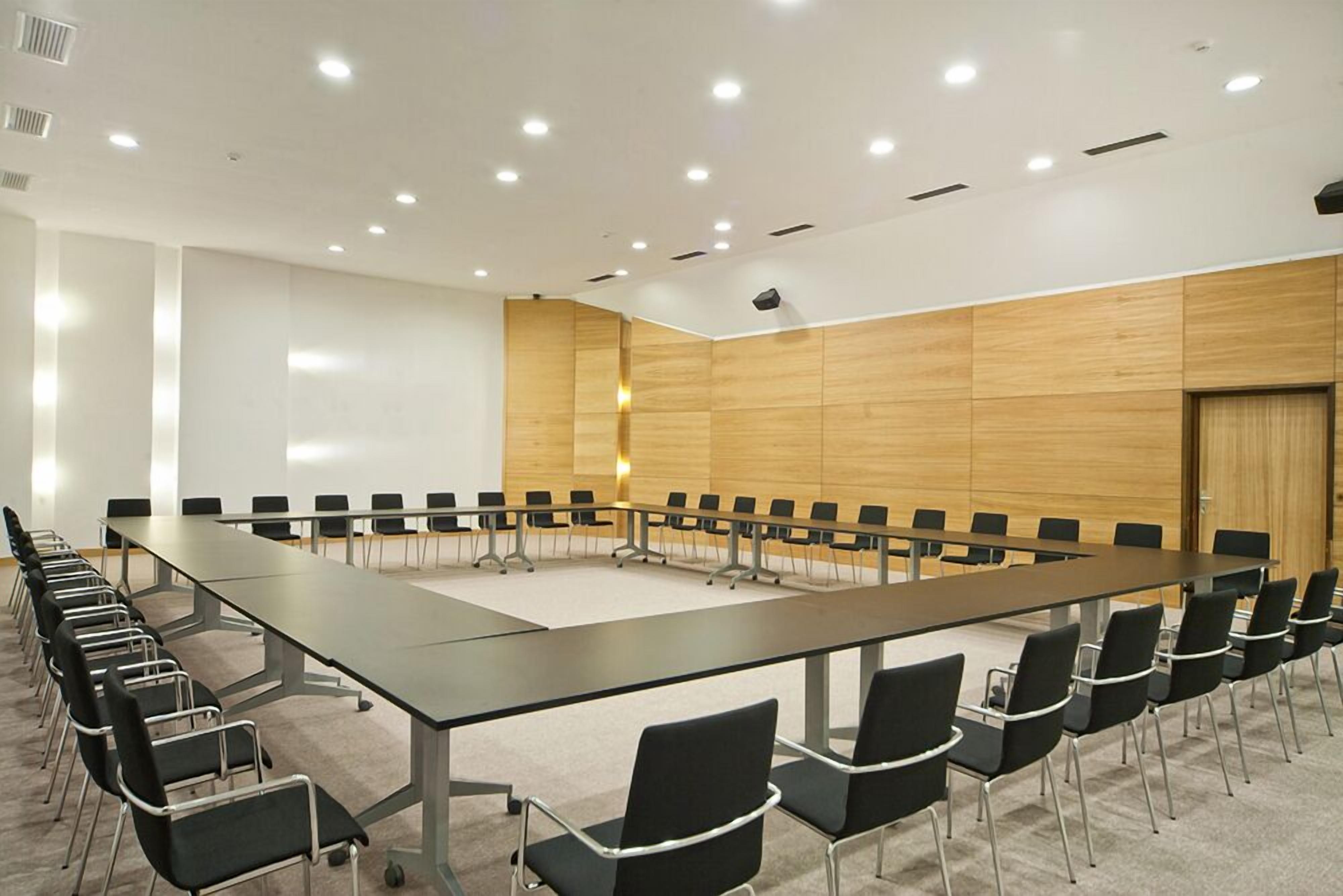 Sala de conferência com uma longa mesa e cadeiras, paredes brancas, painéis de madeira e luminárias embutidas no teto.