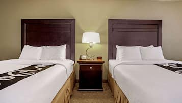 Quarto de hotel com duas camas, cada uma com roupa de cama branca e cabeceira escura. Há uma mesinha de cabeceira entre as camas, com um abajur, telefone e relógio. O quarto possui piso coberto por carpete e paredes bege.