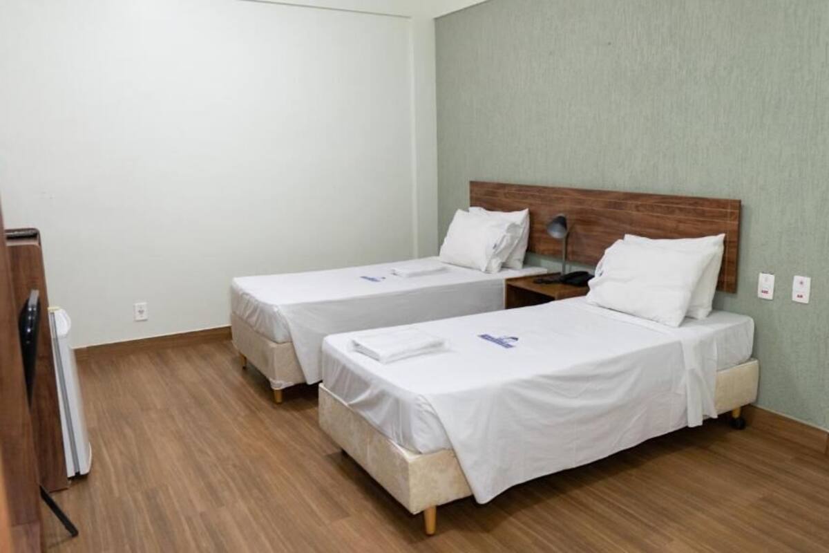 Quarto de hotel com duas camas de solteiro, cabeceira de madeira, mesa de cabeceira com abajur e piso de madeira.
