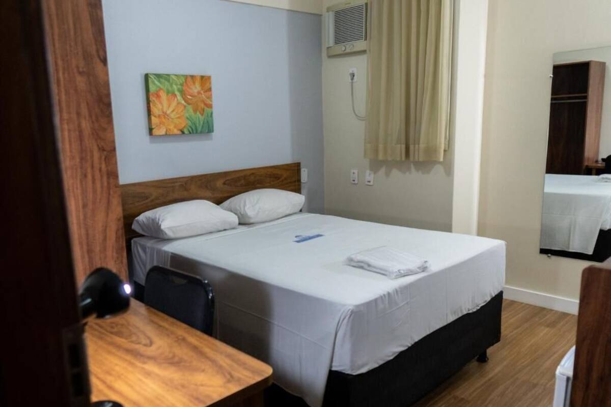 Quarto de hotel com uma cama, uma escrivaninha, uma cadeira, um quadro na parede, um ar condicionado e um guarda-roupa.