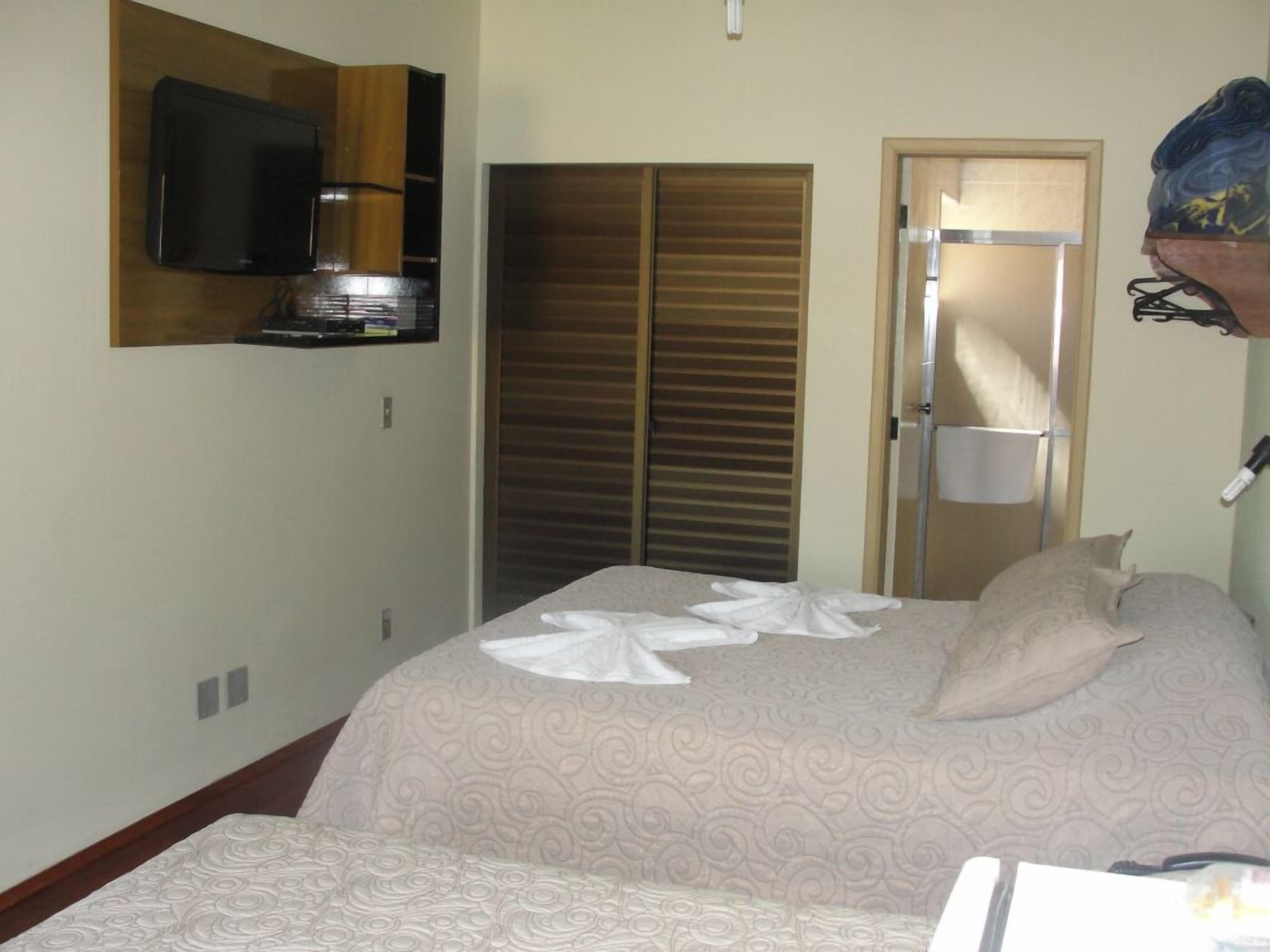 Quarto de hotel com cama, televisão fixada na parede, roupeiro com portas deslizantes e casa de banho visível através de uma porta.