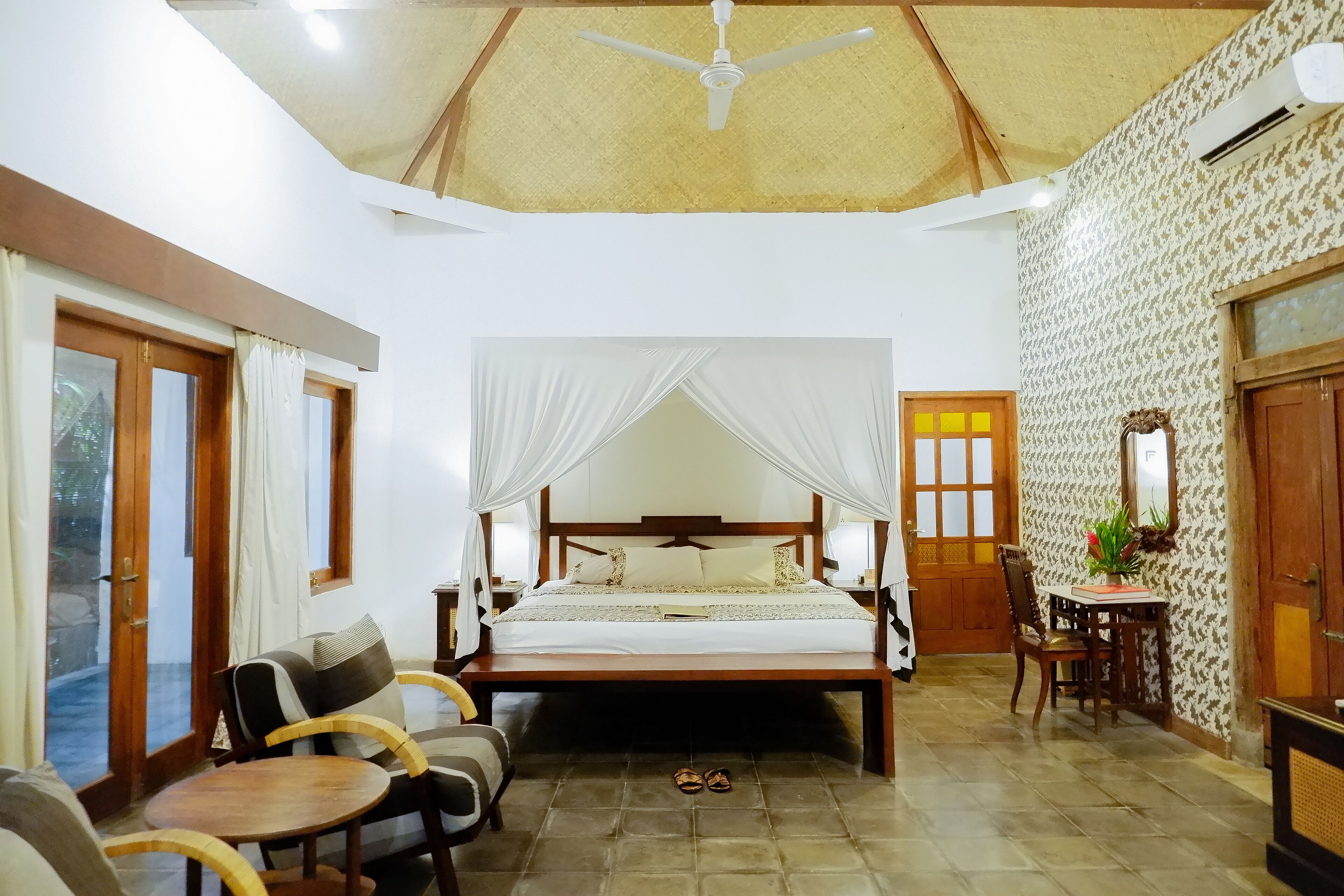 Quarto espaçoso com uma cama grande, um ventilador de teto, uma mesa de jantar com cadeiras e vista através de uma porta de vidro.