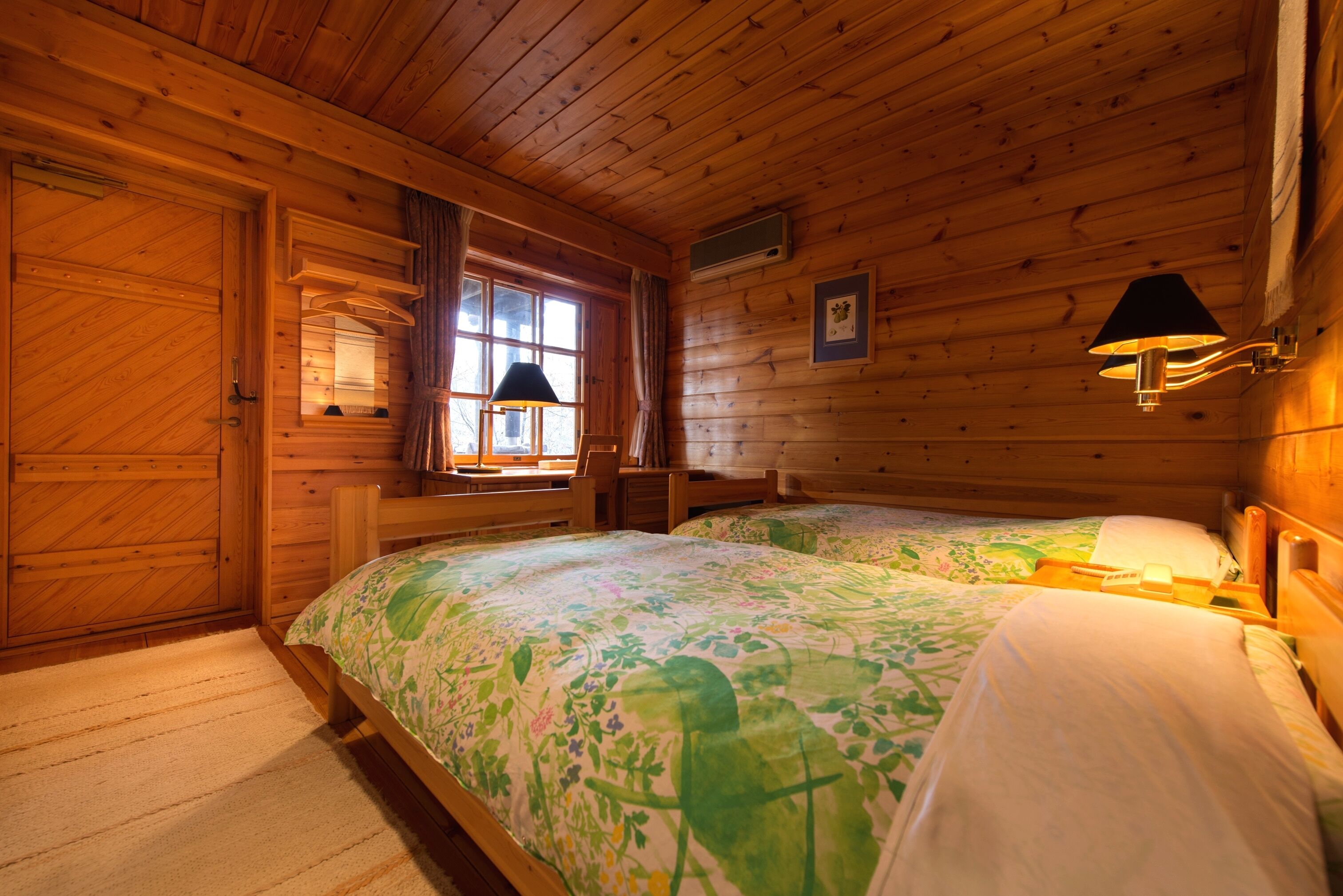 Quarto de cabana de madeira com duas camas, uma janela com cortinas e uma luminária fixada na parede.