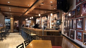 Interior de restaurante com mesas e cadeiras de madeira, área de bar e quadros nas paredes.
