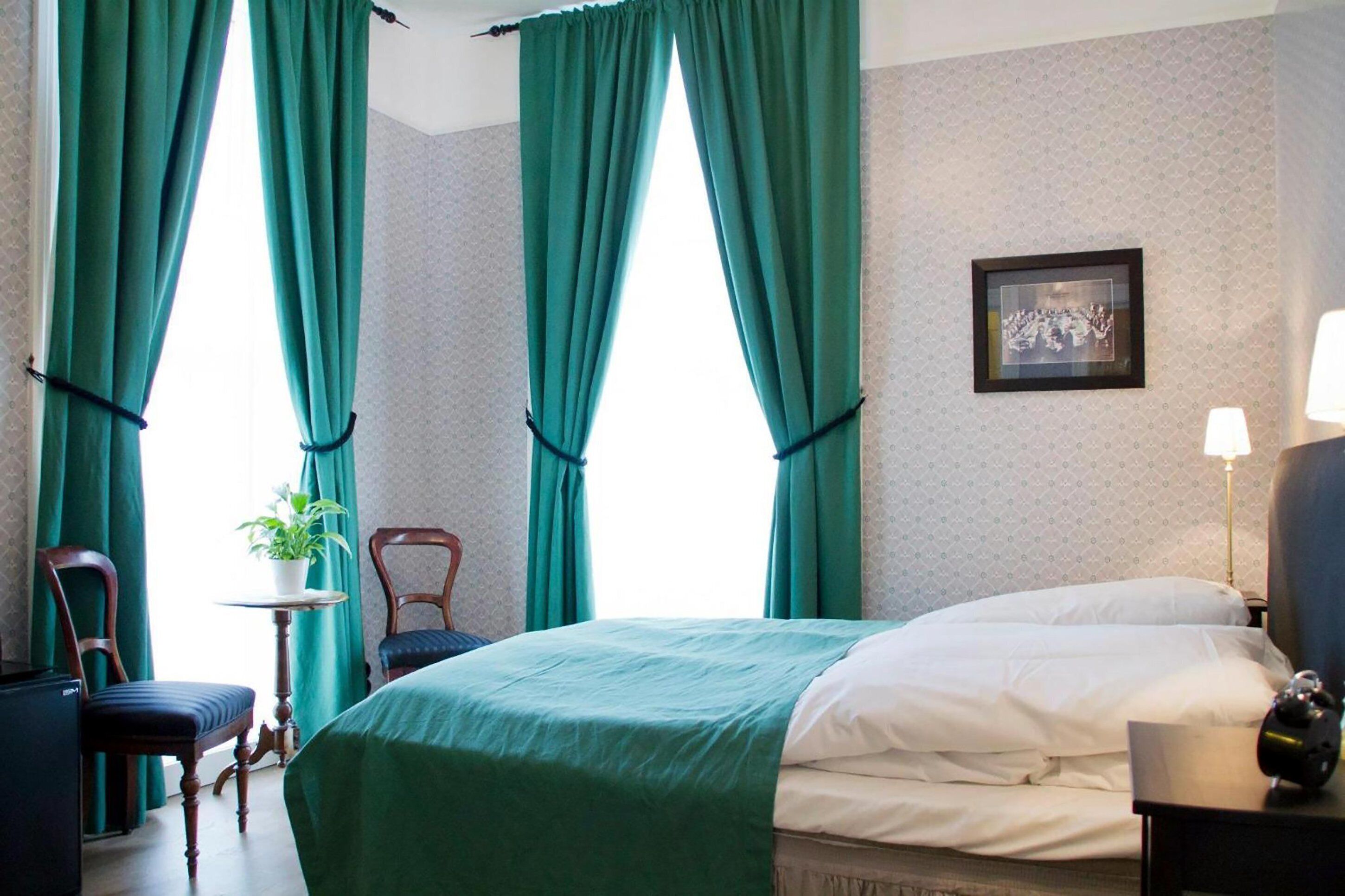 Quarto de hotel com uma cama, duas cadeiras, uma mesa pequena com uma planta, um abajur e um quadro na parede.