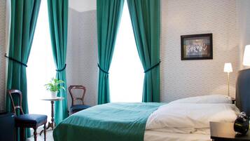 Quarto de hotel com uma cama, duas cadeiras, uma mesa pequena com uma planta, um abajur e um quadro na parede.