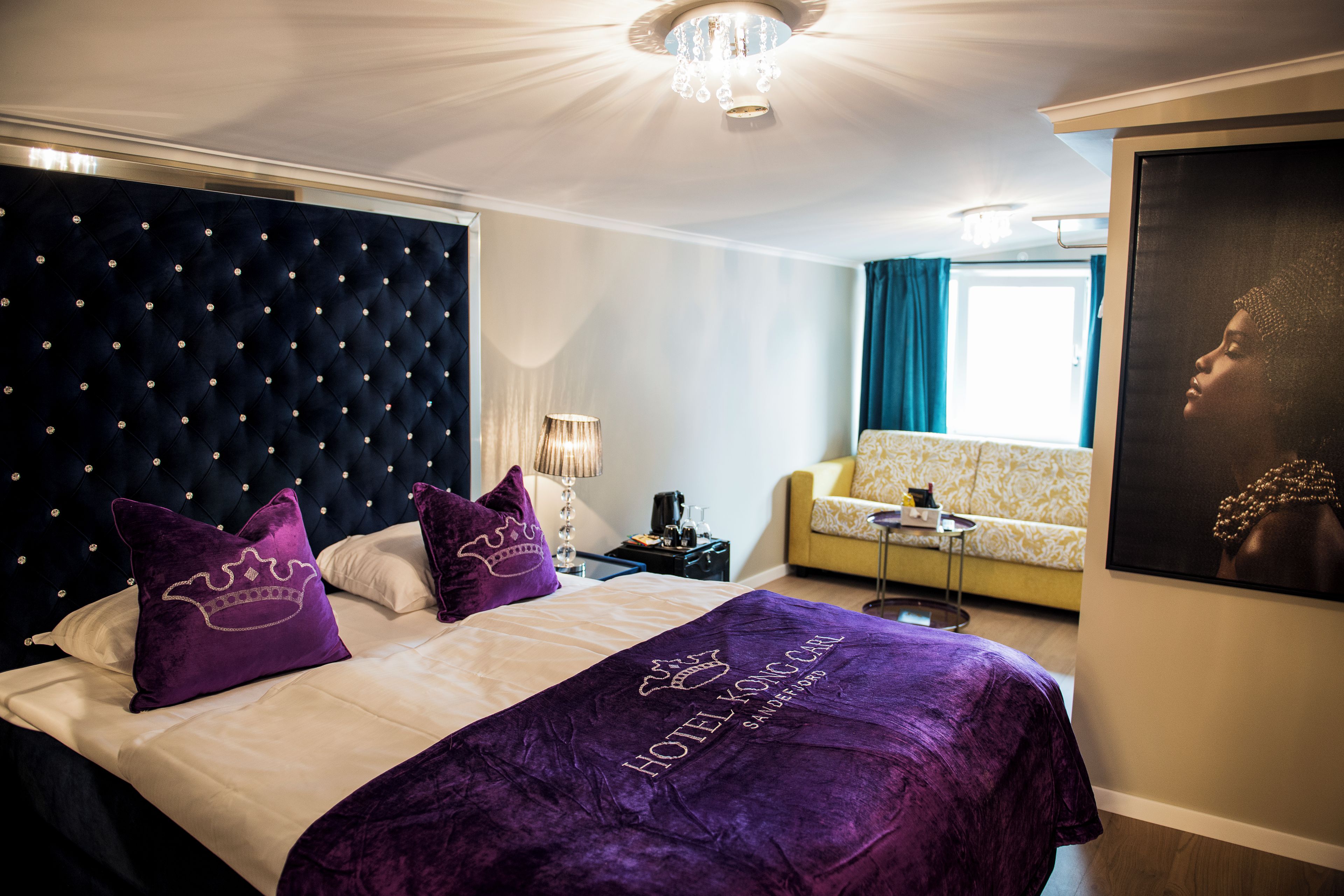 Quarto de hotel com uma cama grande, mesa de cabeceira, luminária, sofá e um quadro na parede.