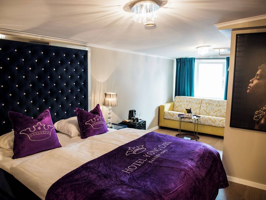 Quarto de hotel com uma cama grande, mesa de cabeceira, luminária, sofá e um quadro na parede.