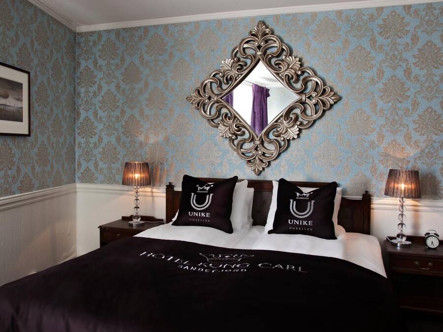 Quarto de hotel com uma cama grande, duas mesas de cabeceira, um espelho com moldura decorativa e parede com papel de parede estampado.