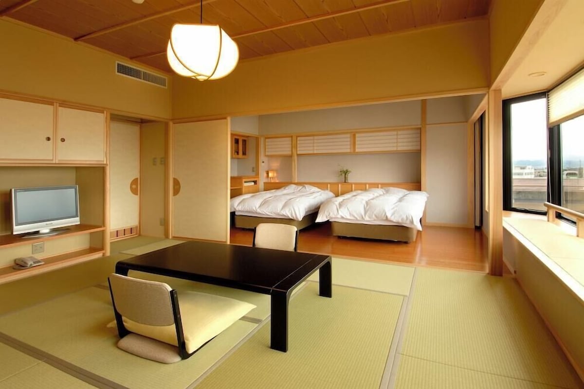 Quarto em estilo japonês tradicional com tatames, uma mesa baixa, televisão e cama com roupa de cama branca.