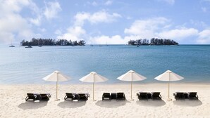 Uma praia com cadeiras de praia e guarda-sóis, mar calmo e barcos ao longe.