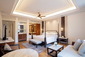 Quarto de hotel moderno com uma cama grande, área de estar com sofá e poltrona, uma pequena mesa e um ventilador de teto.