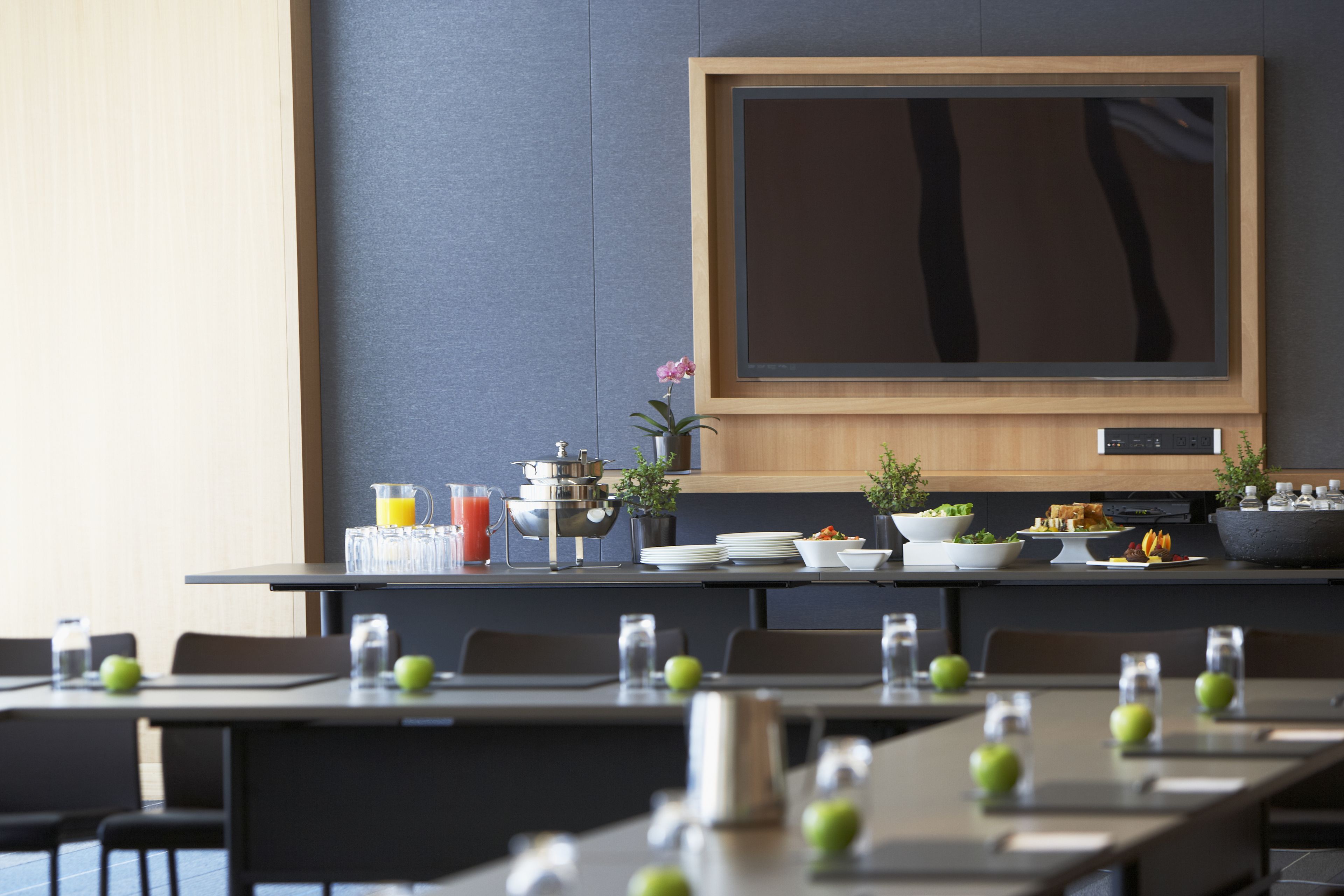 Uma área de refeições moderna com uma TV de tela plana na parede, mesa de buffet com diversos pratos e mesas organizadas com cadeiras.
