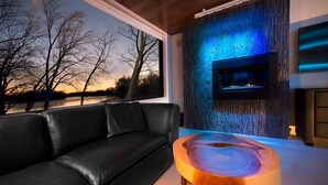 Sala de estar moderna com sofá de couro preto, mesa de centro de madeira, lareira e uma grande janela com vista para árvores e um pôr do sol.