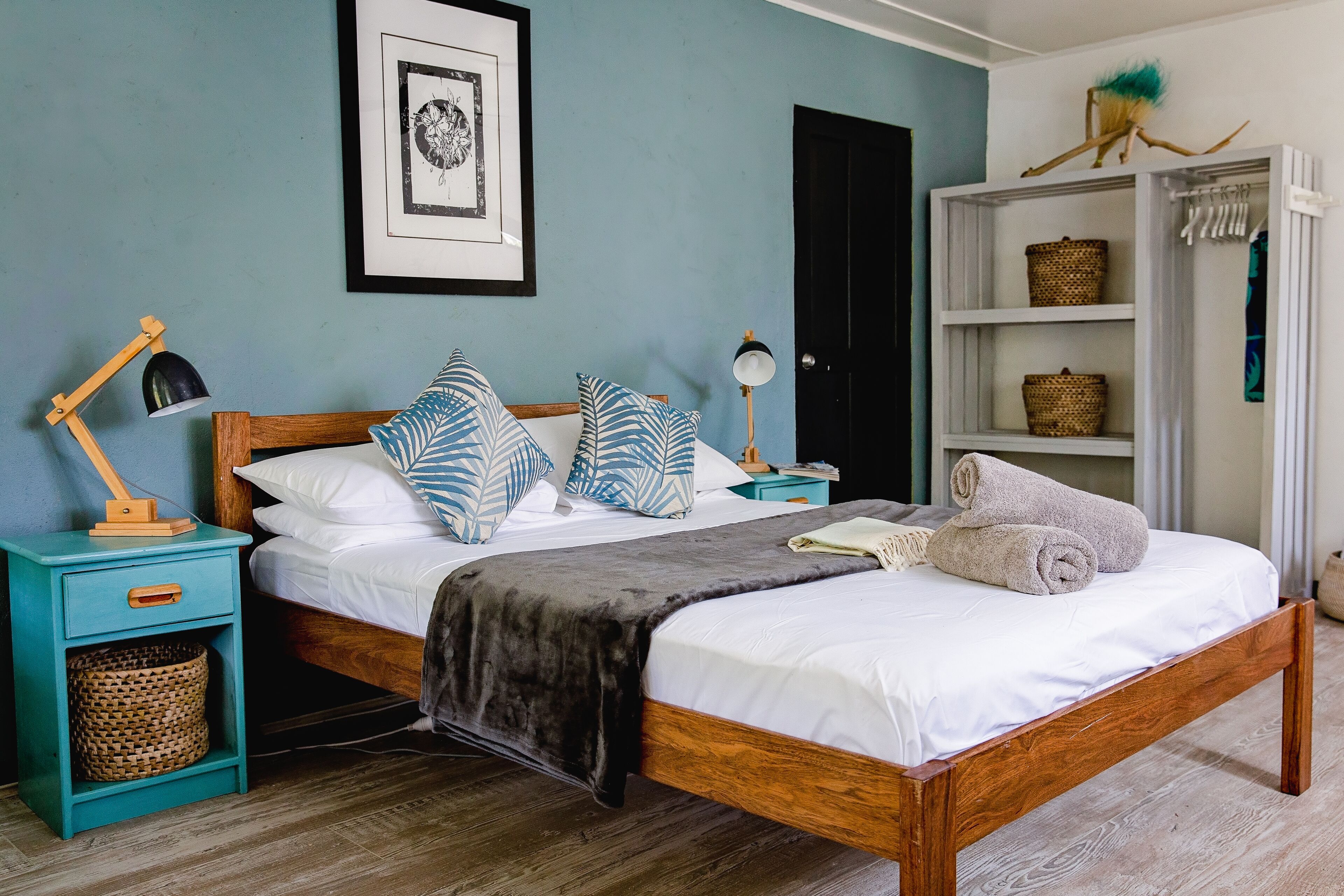 Cama bem arrumada com estrutura de madeira, mesa de cabeceira azul-turquesa com abajur, quadro na parede e guarda-roupa branco com cestos.