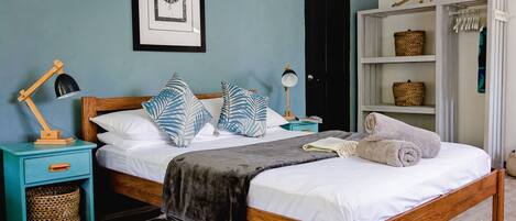 Cama bem arrumada com estrutura de madeira, mesa de cabeceira azul-turquesa com abajur, quadro na parede e guarda-roupa branco com cestos.