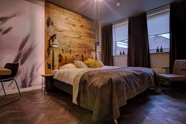 Quarto moderno com cabeceira de madeira, cama com cobertor cinza, mesa de cabeceira, uma cadeira e janela com cortinas.