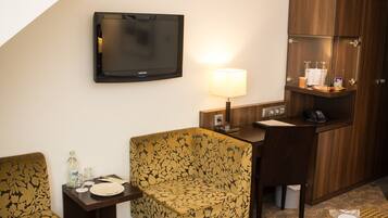 Quarto de hotel com sofá, uma mesinha com abajur, televisão, minibar e área de banheiro.