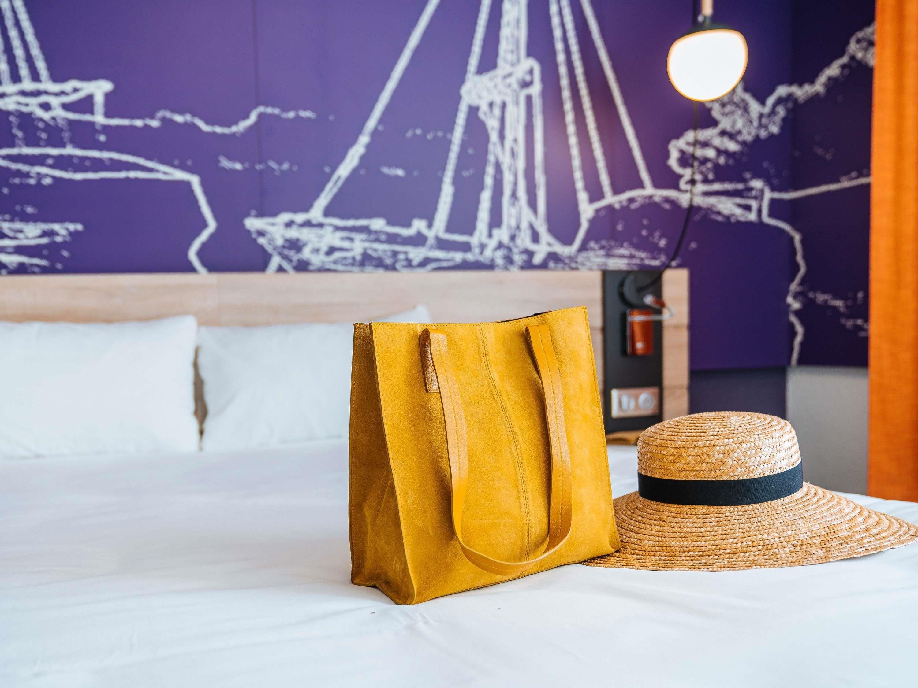 Uma cama com roupa de cama branca, uma sacola amarela mostarda e um chapéu de palha sobre uma superfície branca.