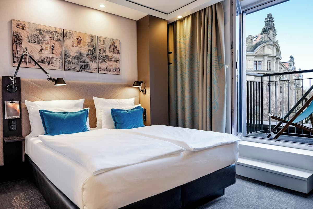 Quarto de hotel com uma cama grande, duas luminárias de cabeceira, uma mesa de cabeceira e uma janela com vista para um edifício.
