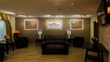 Quarto de hotel com sofá de couro, duas poltronas, uma mesinha e duas luminárias. Há bandeiras americanas nas paredes e uma placa da Hawthorn Suites by Wyndham.