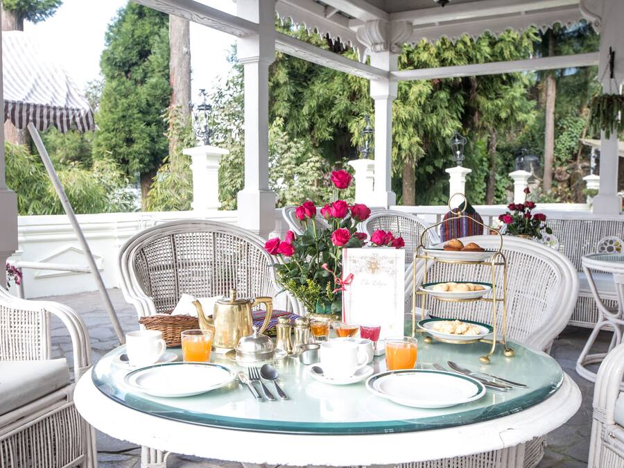 Mesa de vidro redonda posta para o café da manhã em área externa coberta, com cadeiras de vime, bandeja escalonada de doces e um vaso de rosas vermelhas.