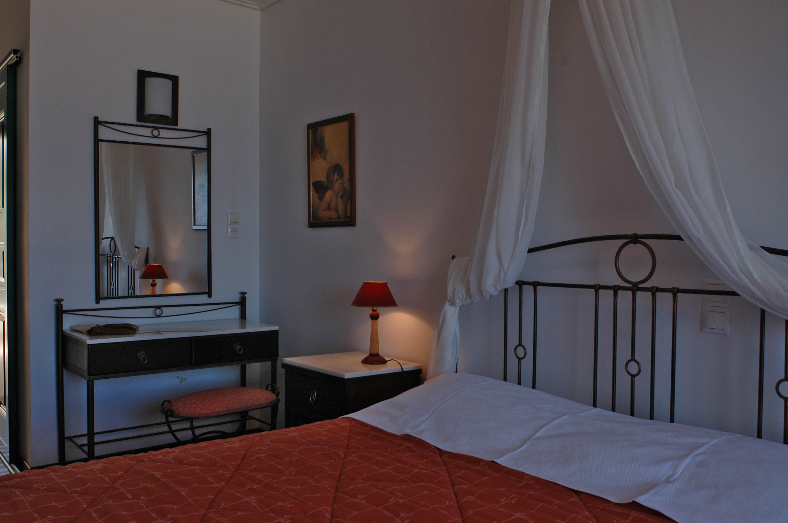 Um quarto com cama, mesa de cabeceira, escrivaninha, cadeira, espelho e um quadro na parede.