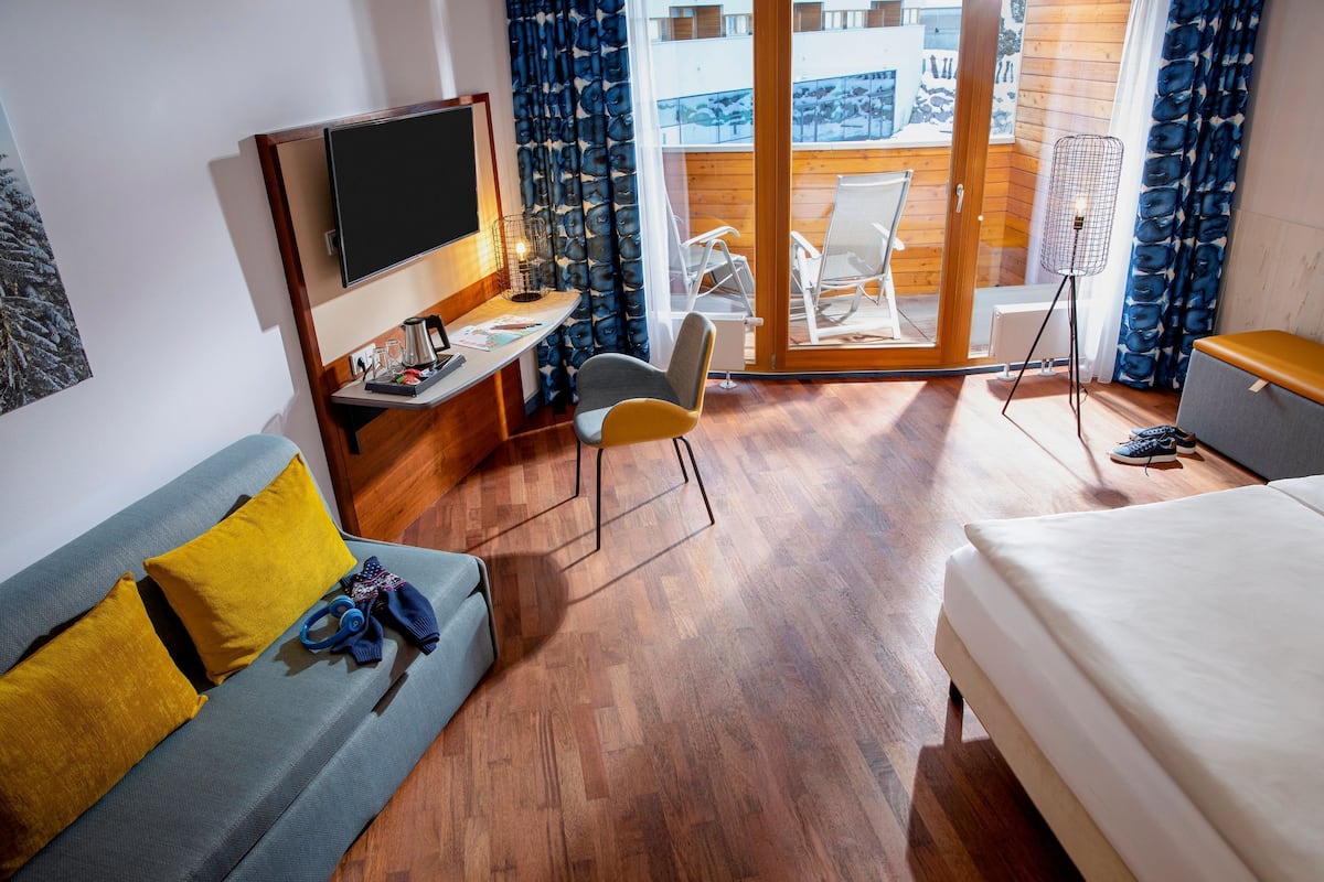 Quarto de hotel com cama, sofá com almofadas amarelas, escrivaninha com televisão e uma cadeira.