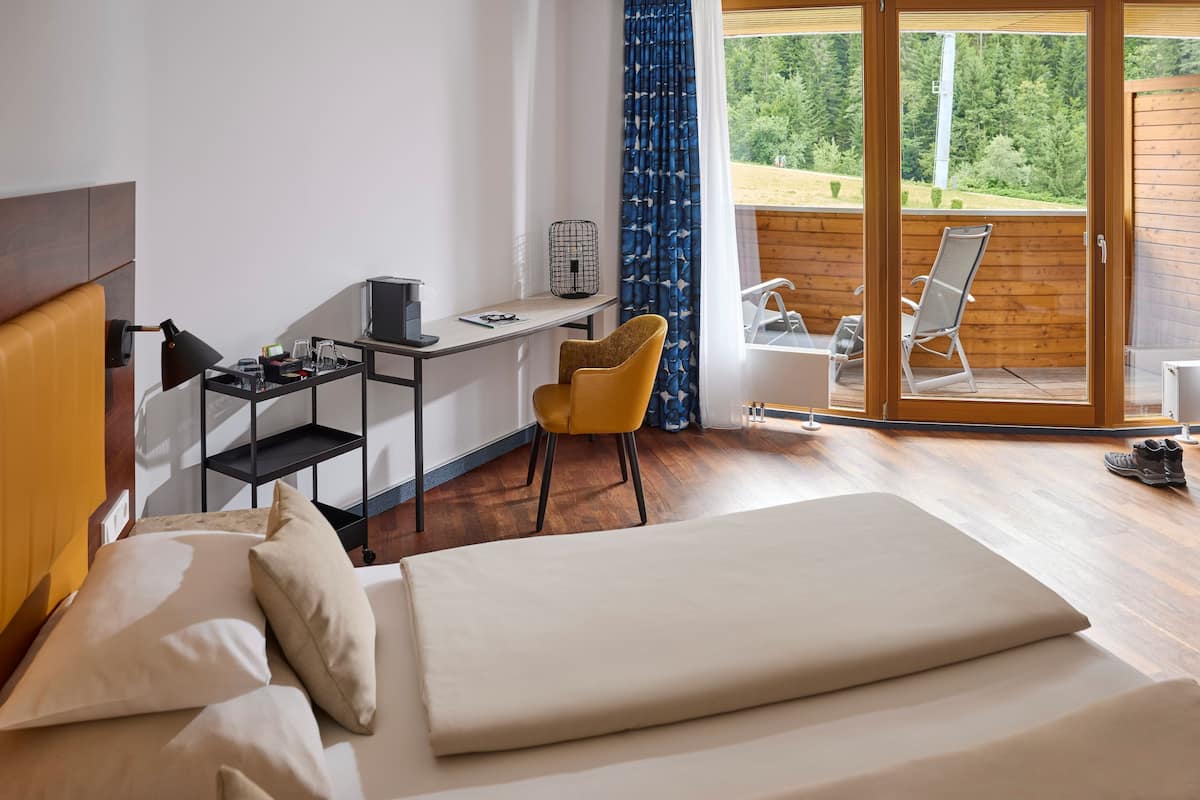 Quarto com cama, escrivaninha com computador, cadeira e vista para uma varanda com mobiliário exterior.