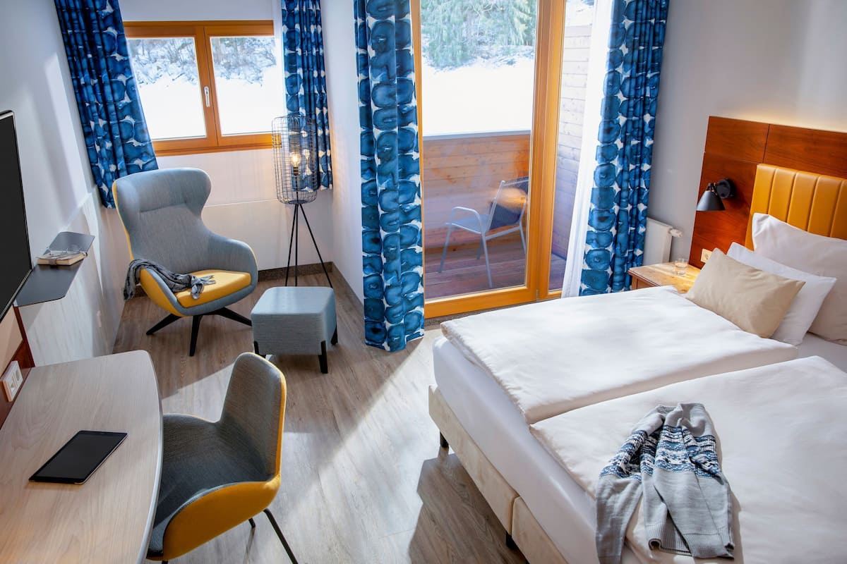 Quarto de hotel com cama, escrivaninha, cadeira e janela com cortinas.