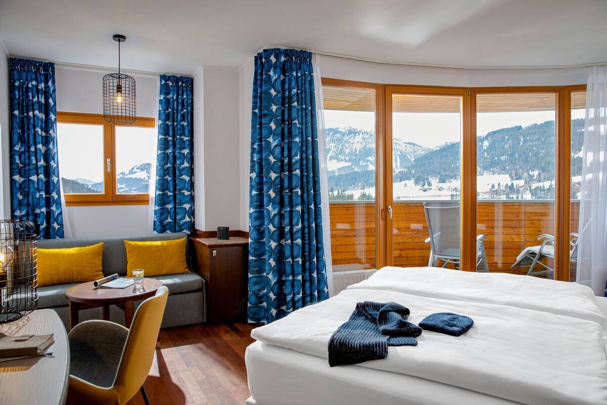 Quarto de hotel com cama, sofá, escrivaninha e vista para montanhas.