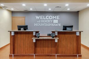 Uma mesa de recepção com o letreiro "WELCOME THE ROCKY MOUNTAIN STATE". Há dois monitores de computador e uma placa "Hilton Honors" sobre a mesa.