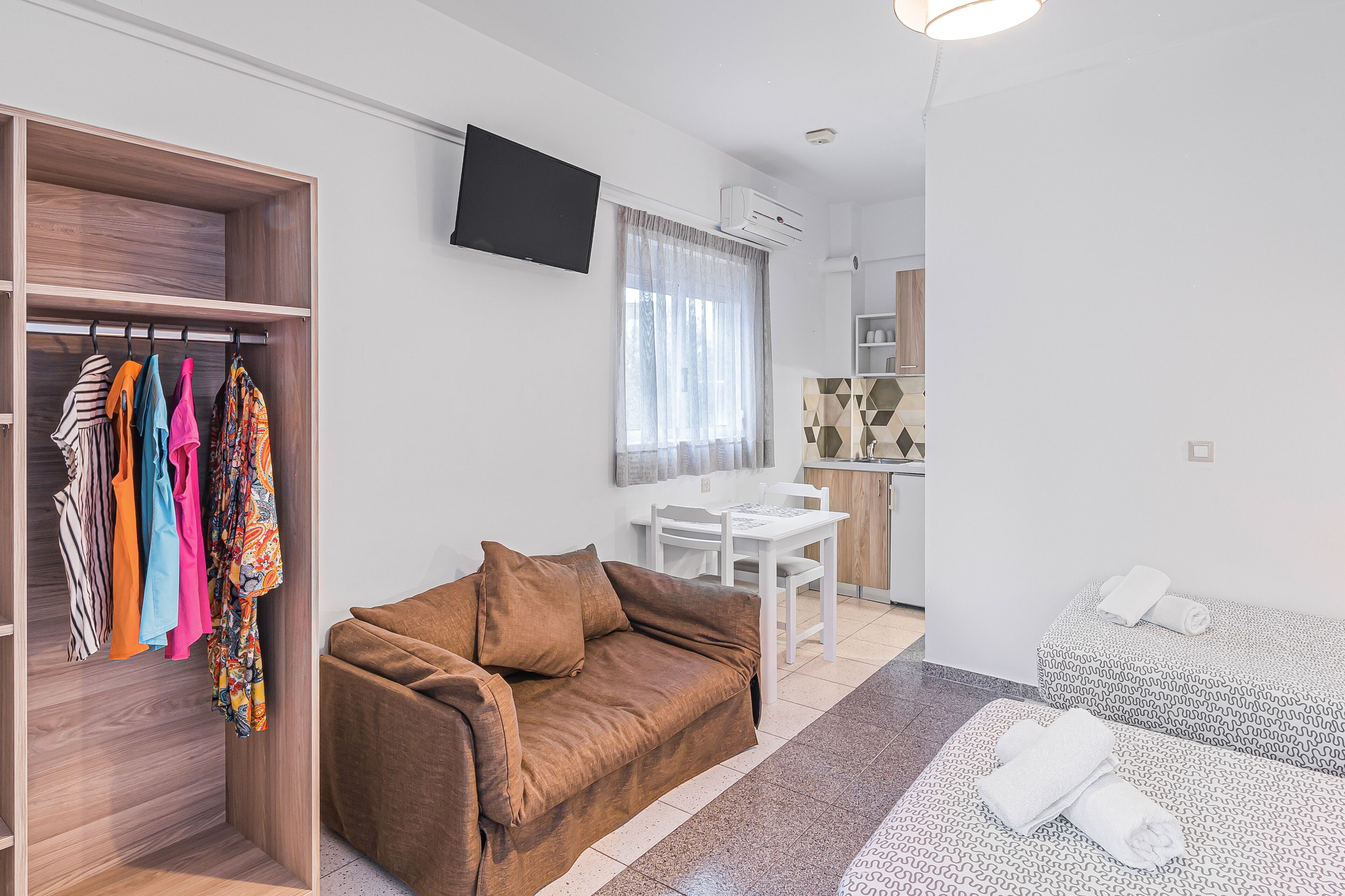 Quarto de hotel moderno com sofá marrom, mesa de jantar, cozinha compacta e TV na parede.