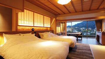 Quarto de hotel com duas camas grandes, teto de madeira, uma janela ampla com vista para uma montanha e varanda com mesa e cadeiras.