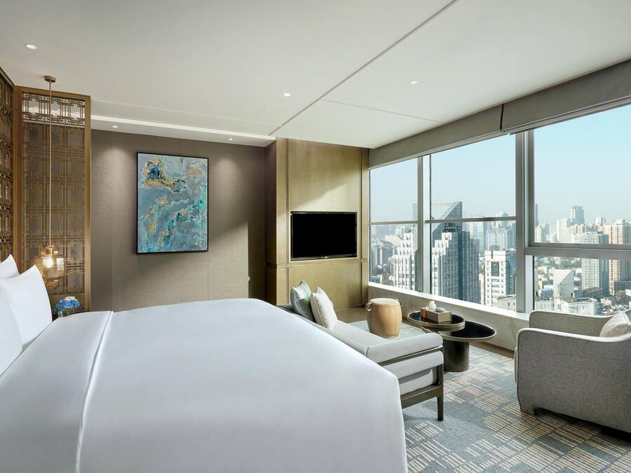 Quarto de hotel com uma cama grande, televisão de tela plana, vista para a cidade, um biombo decorativo e uma pequena mesa com um vaso.