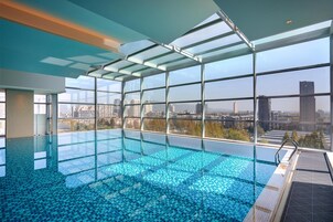 Piscina infinita em ambiente interno, com azulejos azuis cristalinos e teto de vidro, proporcionando vista para a cidade.